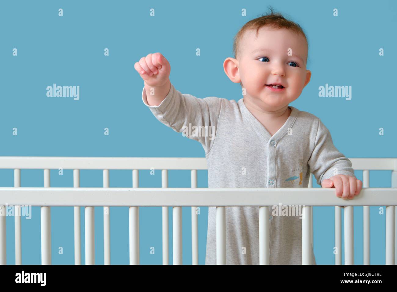 Happy infant baby boy with raised hand gesture in crib, studio blue ...
