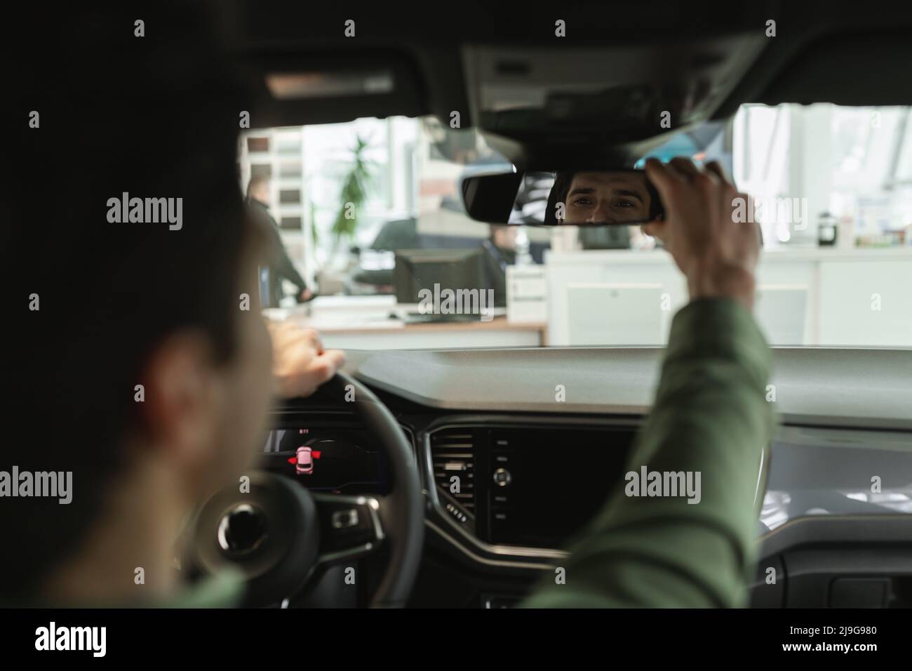 Happy young guy sitting in new car, adjusting rear view mirror ...