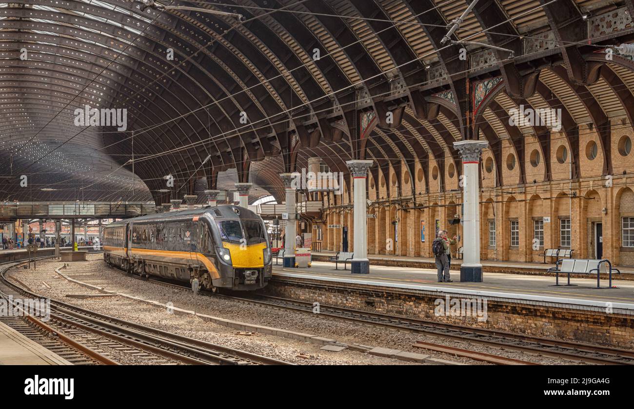 A modern train stands by a historic platform with columns and under an ...