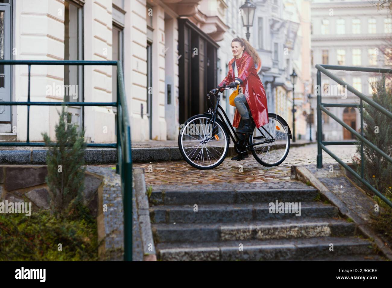 happy modern woman in red rain coat outside in the city riding bicycle ...