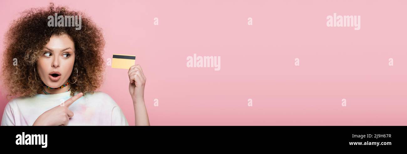 Curly woman pointing at credit card with finger isolated on pink ...