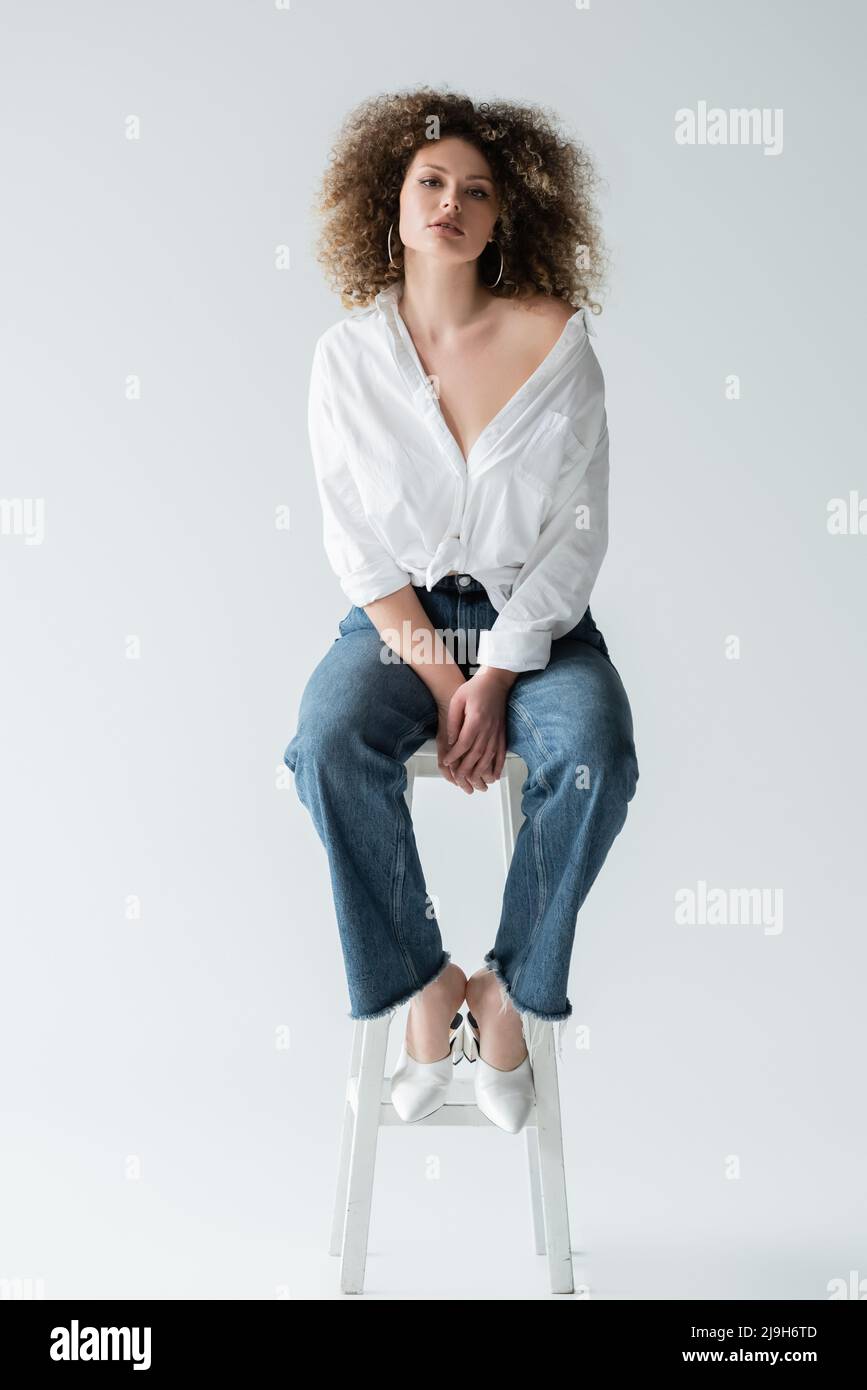Stylish curly woman in blouse sitting on chair on white background ...
