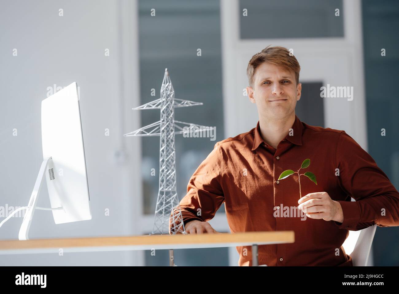 Smiling engineer holding leaf sitting by electricity pylon model at ...