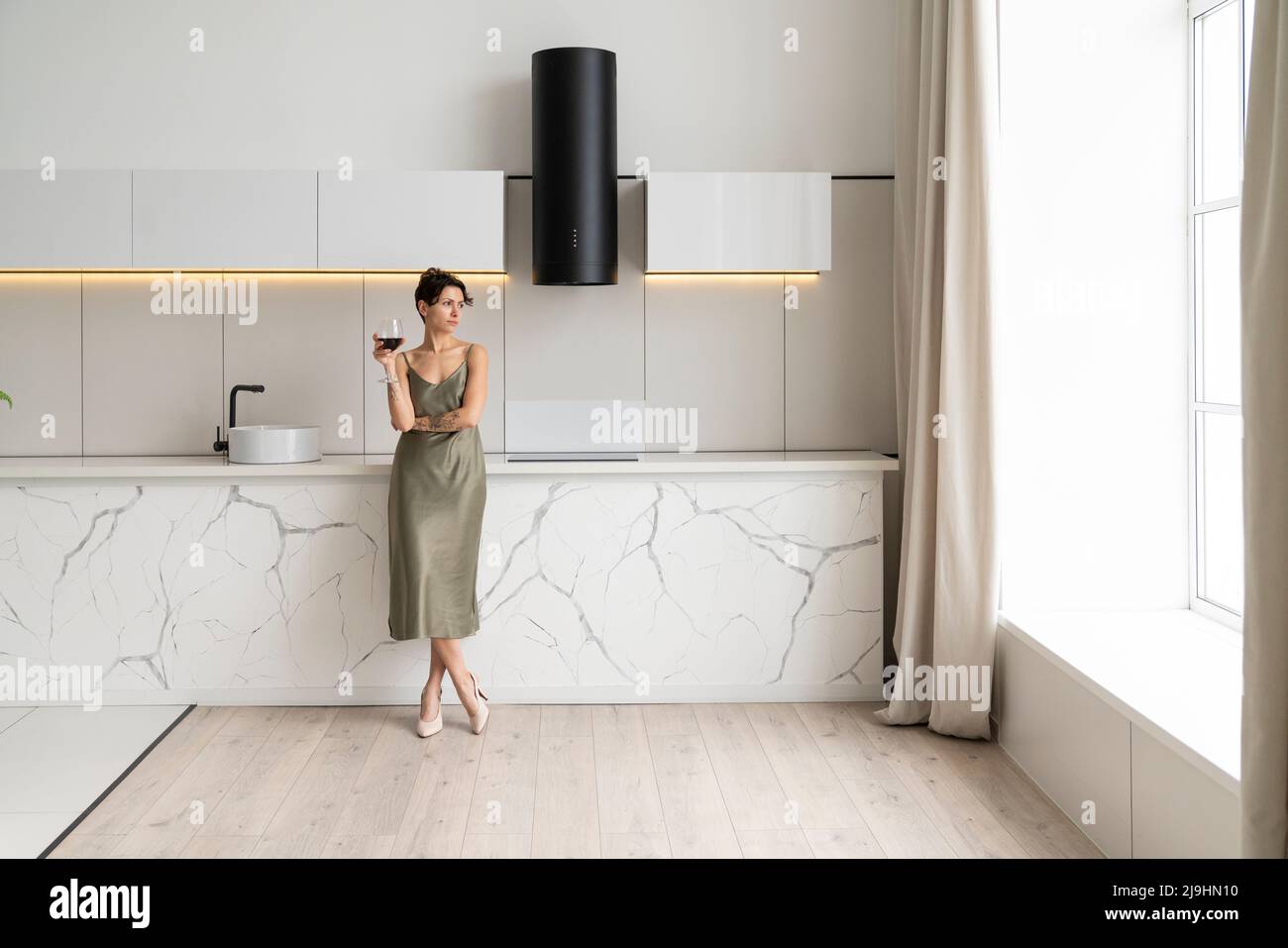 Thoughtful woman holding wineglass looking through kitchen window at ...