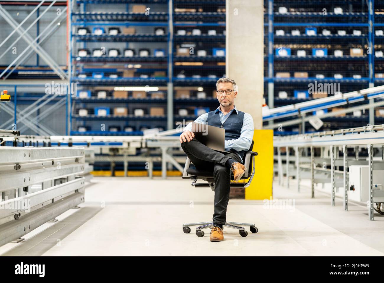 Mature businessman with laptop sitting on chair in warehouse Stock ...