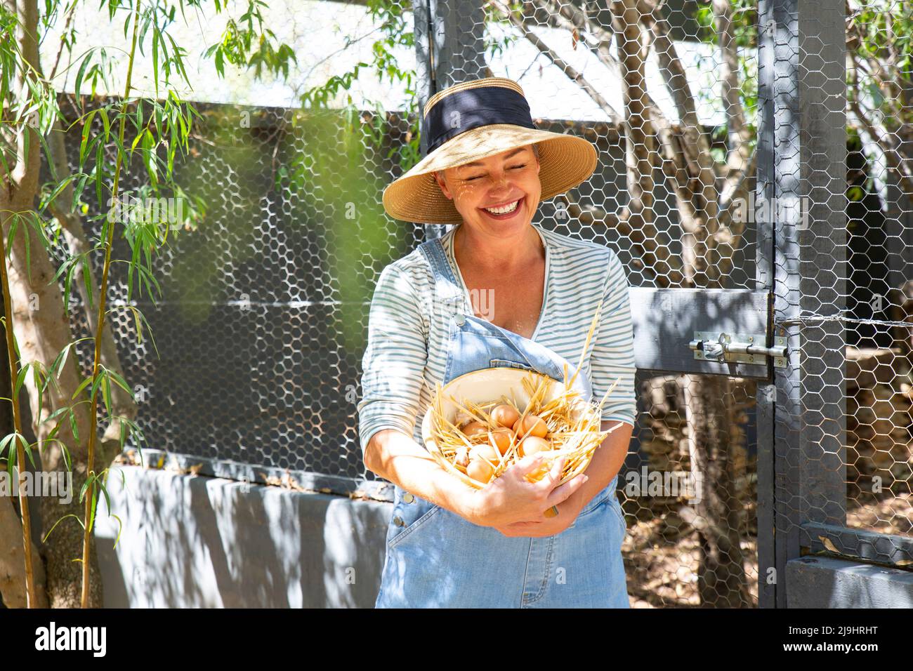 Happy mature farm owner holding egg's bowl in front gate on sunny day ...