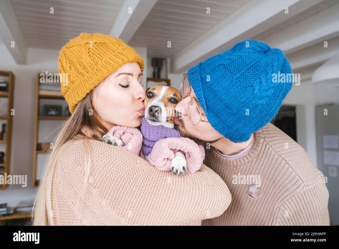 Couple wearing wooly hat at home kissng dog with scarf Stock Photo - Alamy