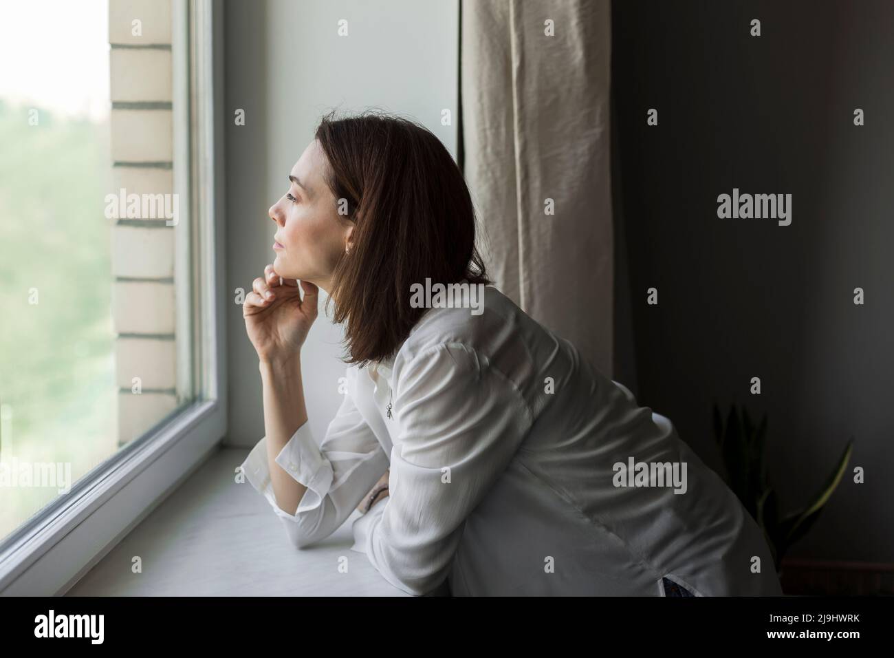 Mature woman with hand on chin looking through window at home Stock ...
