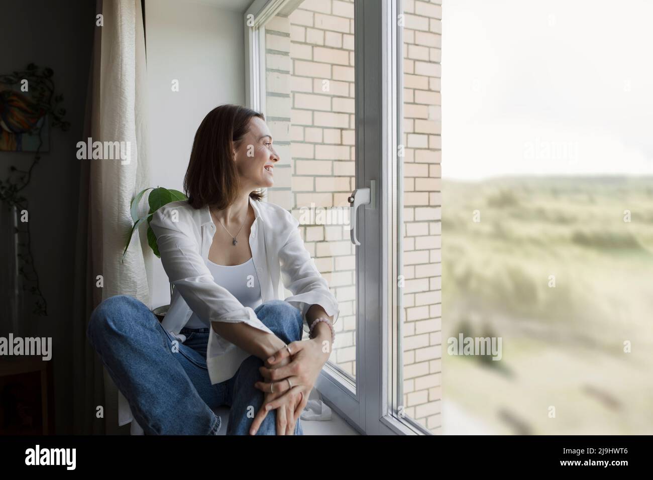 Smiling mature woman sitting on window sill at home Stock Photo - Alamy