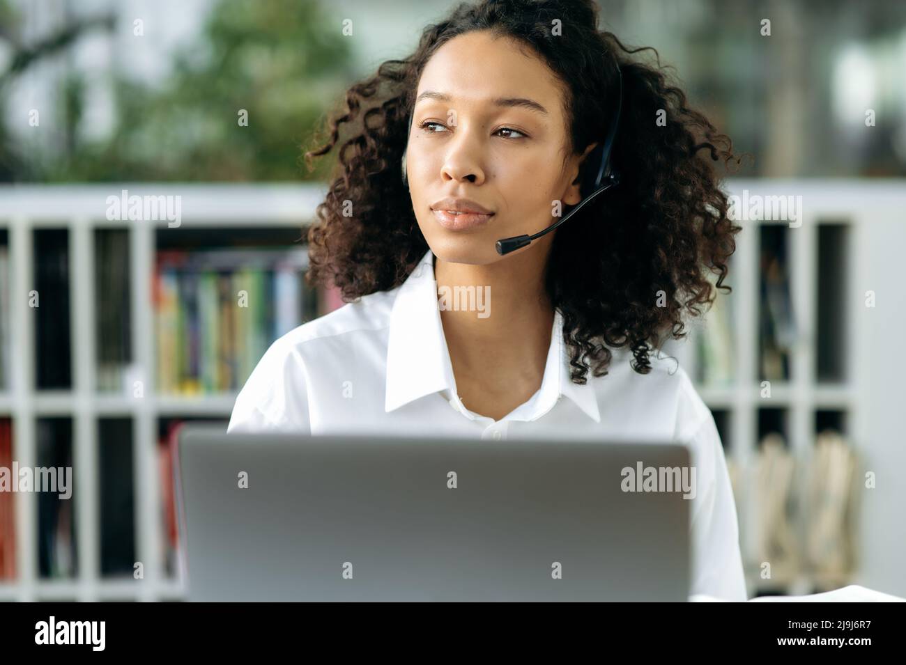 Pensive dark-skinned curly girl in white shirt, call center operator ...