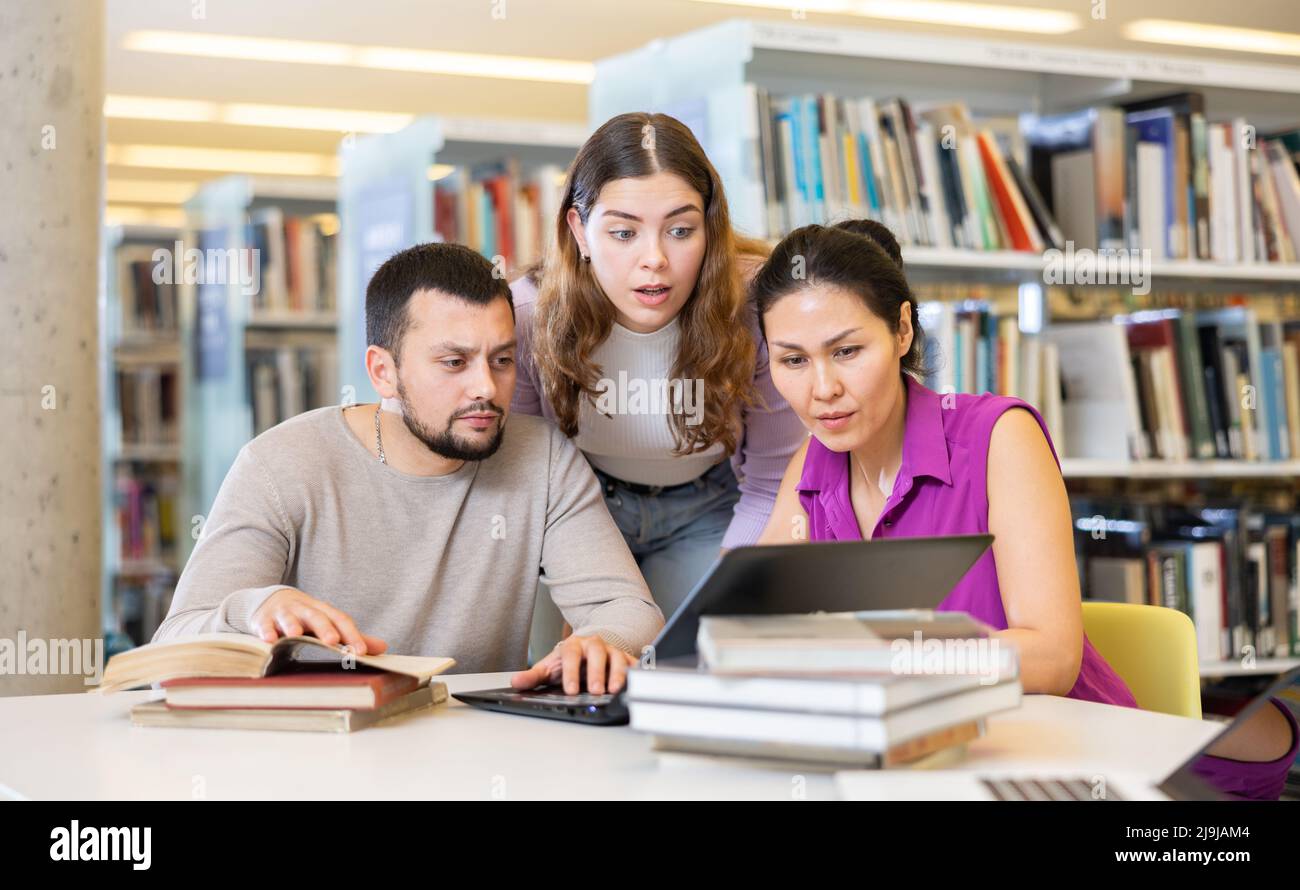 Three adult students work on laptop and read books in a public library ...
