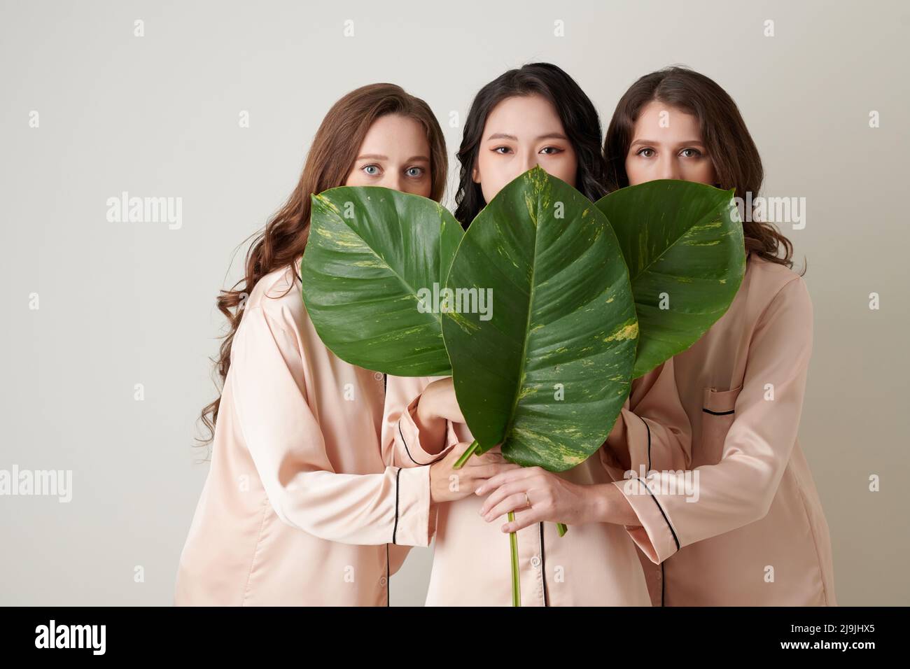 Group of young women in satin pajamas covering lips with big leaves ...