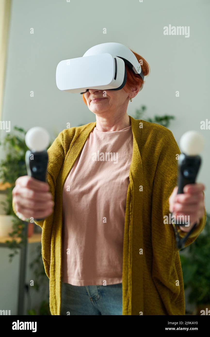 Senior woman in casual clothing using wireless goggles and joysticks to ...