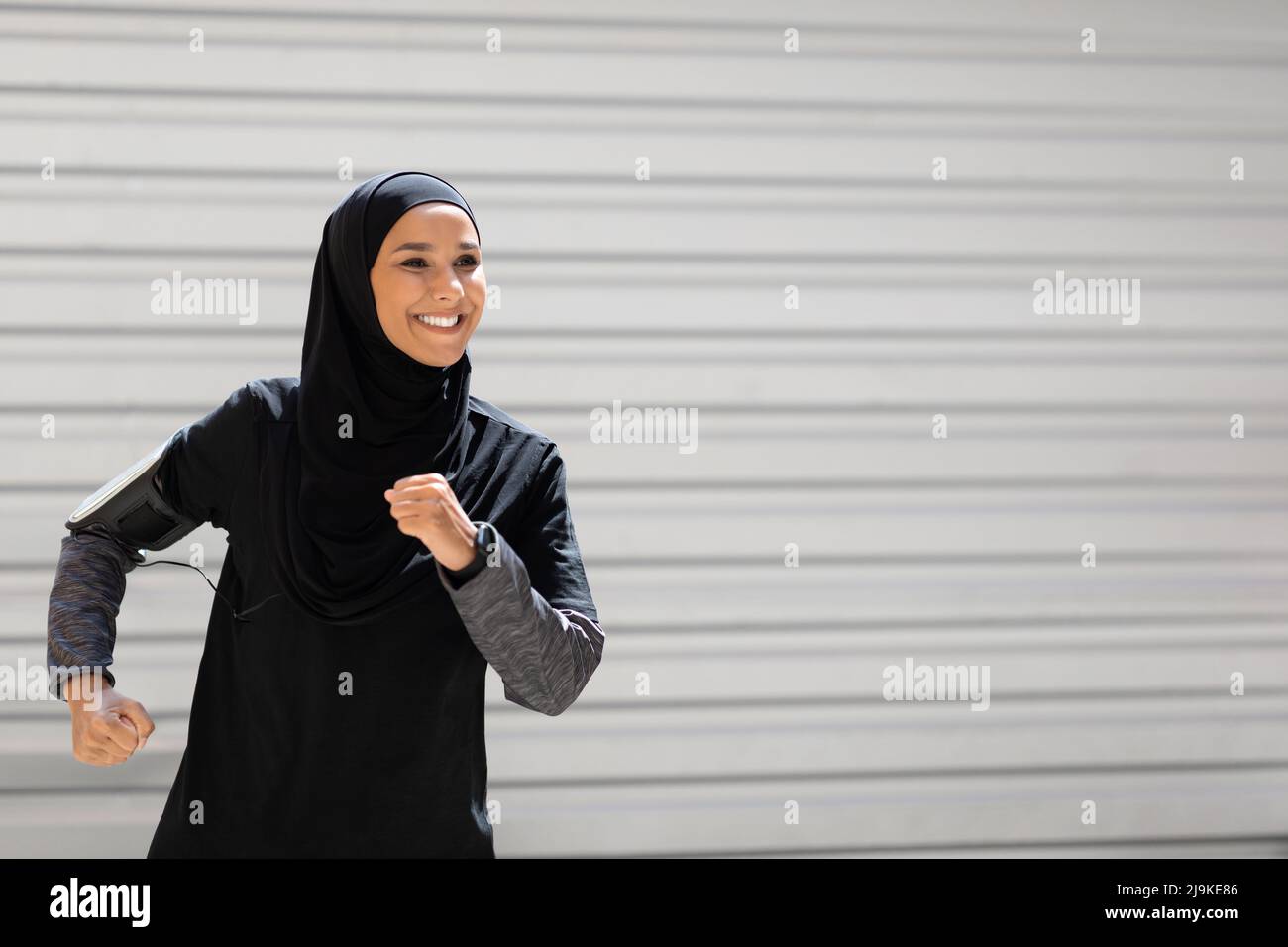 Cheerful young middle eastern female in hijab with phone on her shoulder runs for weight loss ...
