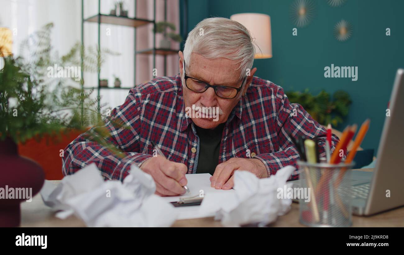 Senior man working on laptop computer, making notes on sheet of paper ...