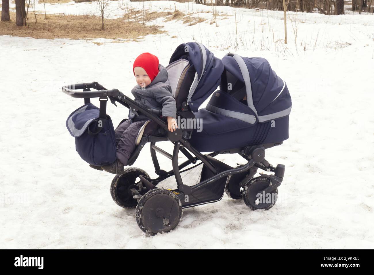 Small child in red cap in double blue pram looks inside and smiles ...