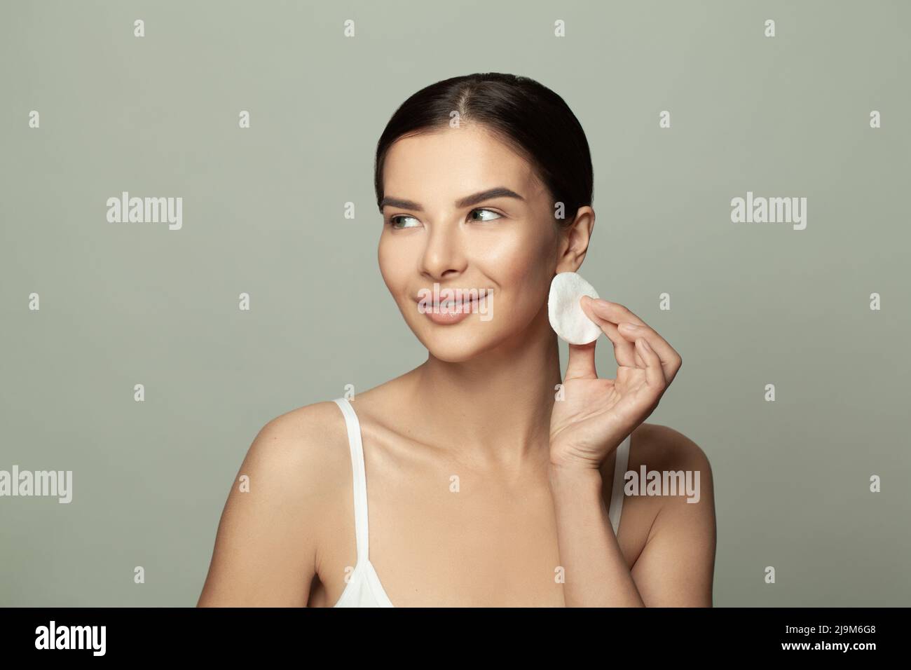 Smiling attractive woman cleaning her face with a cotton pad on gray ...