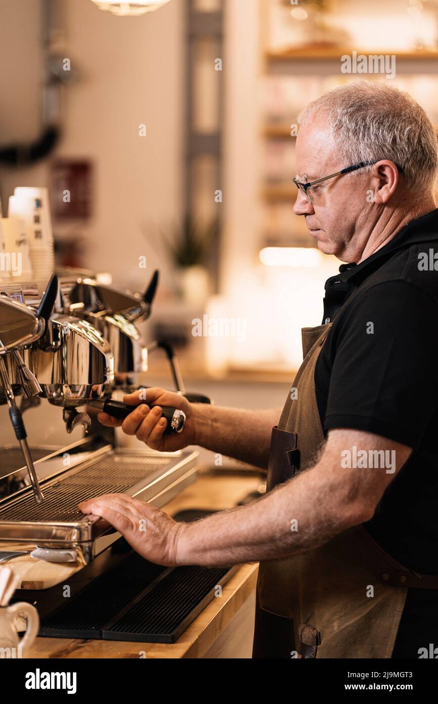 Side view of focused senior male barista in apron and eyeglasses ...