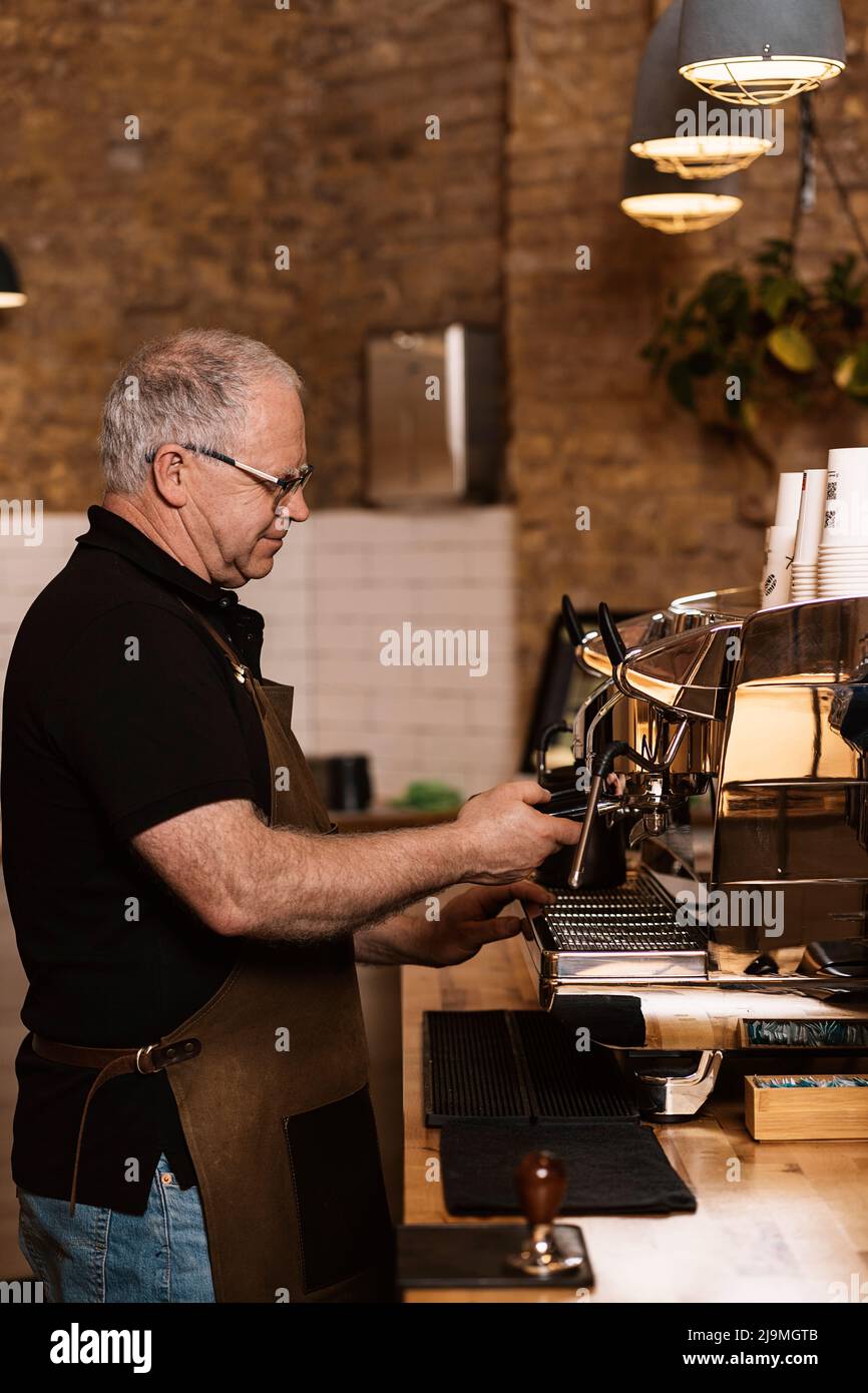 Side view of senior male barista in apron and eyeglasses putting ...