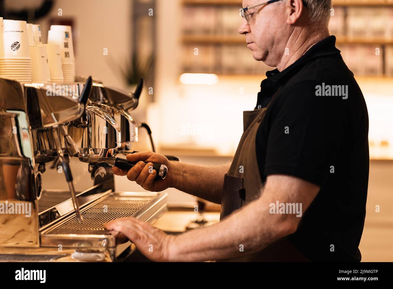 Side view of focused senior male barista in apron and eyeglasses ...