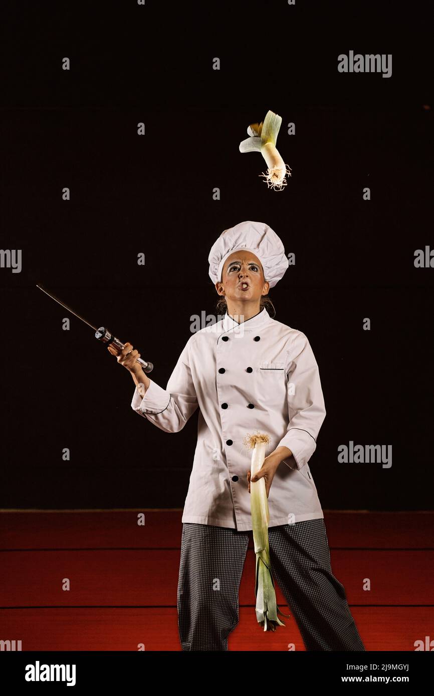 Young female mime artist with makeup in chef uniform and hat making face and cutting fresh leek ...