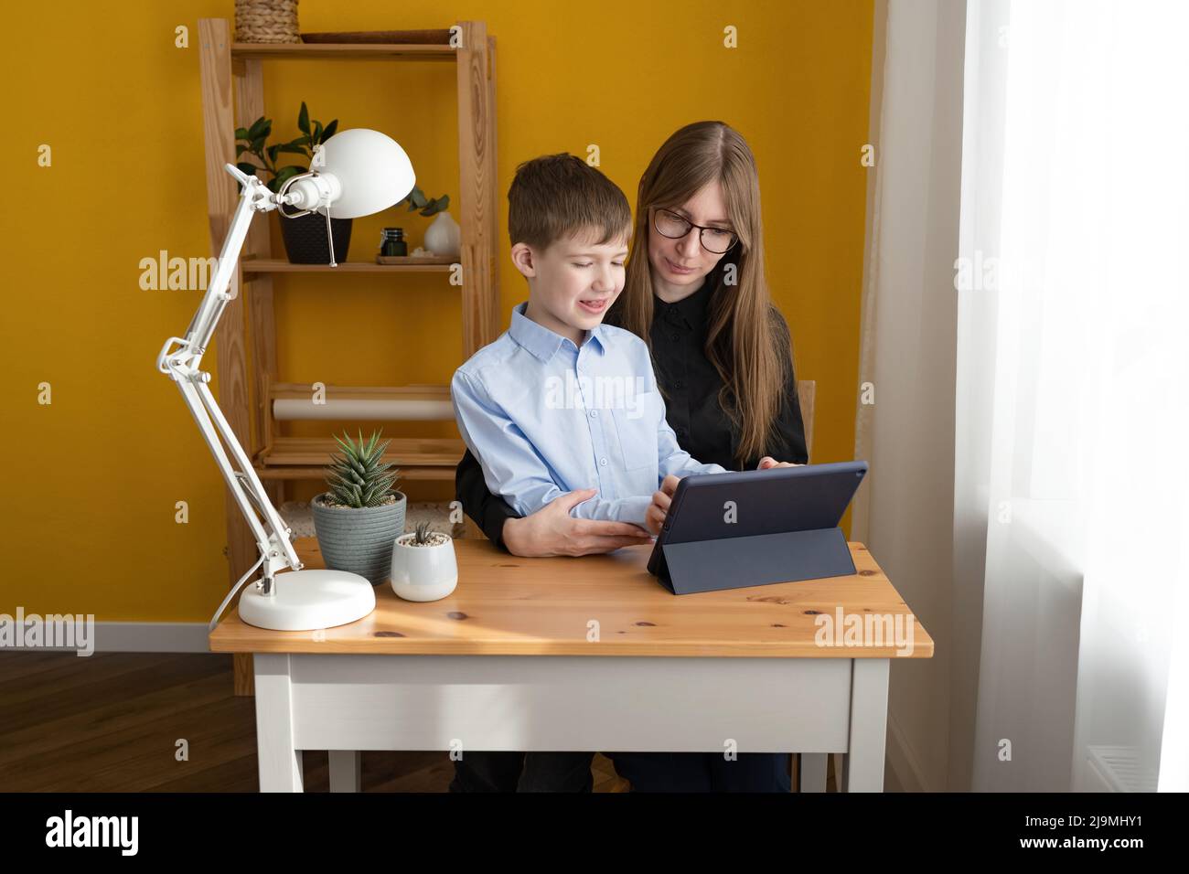 Child in blue shirt browsing tablet with mom at table during remote ...