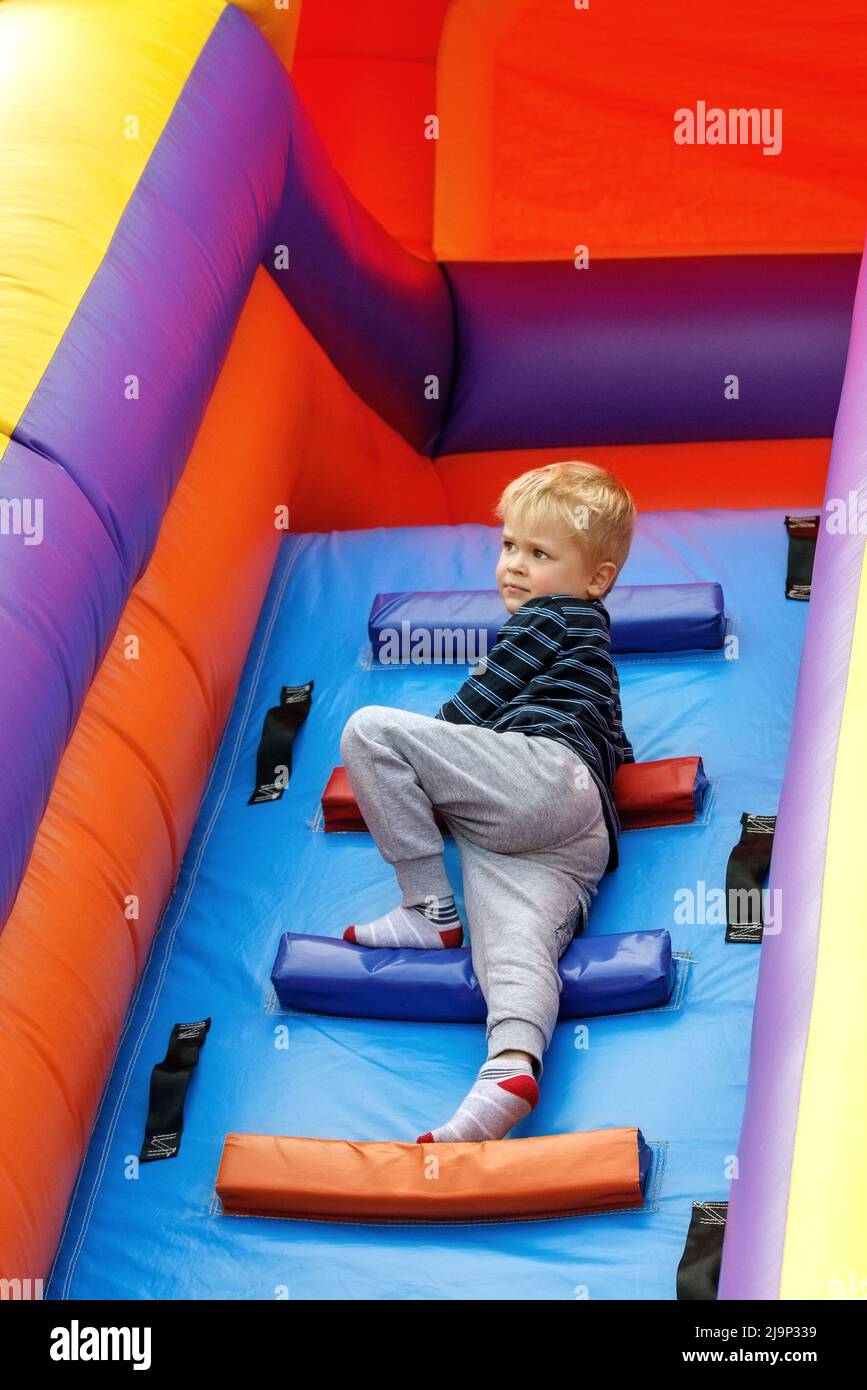 A cute boy climbs a colorful ladder on a high inflatable trampoline and ...
