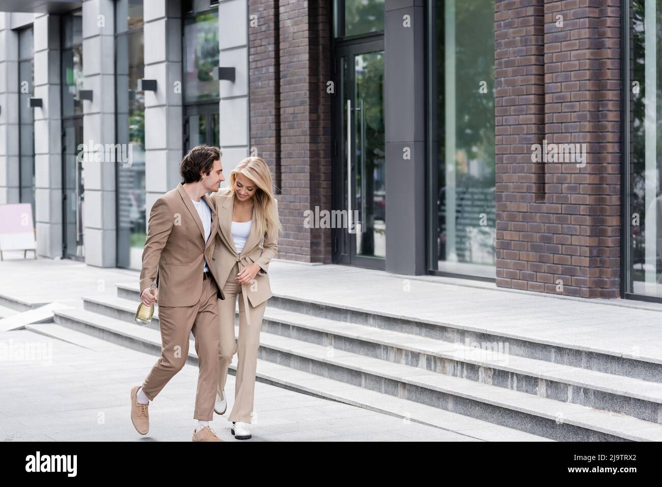 full length of smiling couple in stylish suits walking with champagne ...