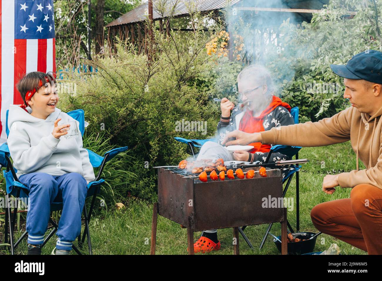 Happy young family make barbecue together in garden. People barbecuing meat on grill. Dad, son ...