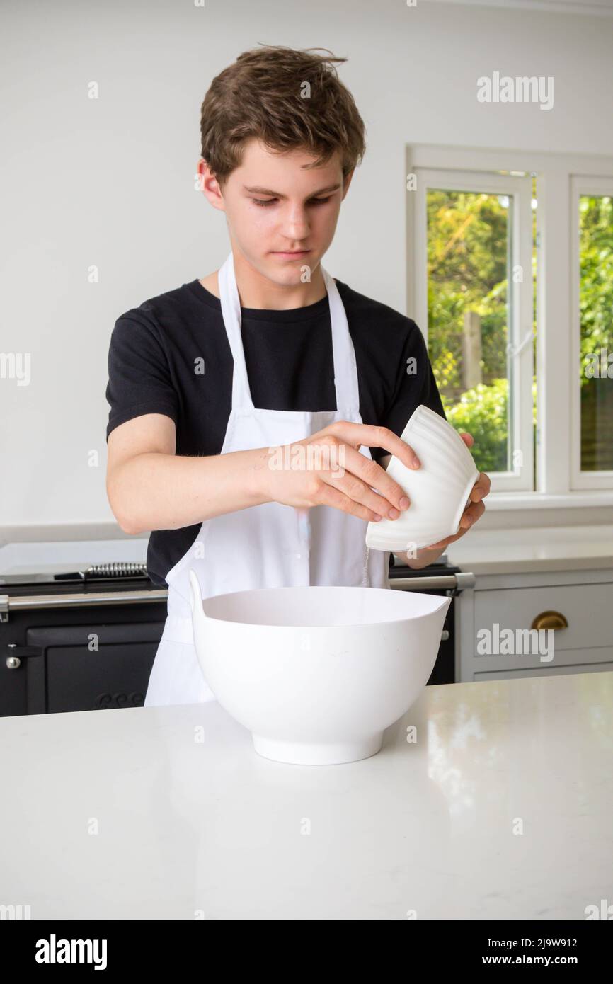 A teenage boy in a domestic kitchen adding ingredients to a mixing bowl ...