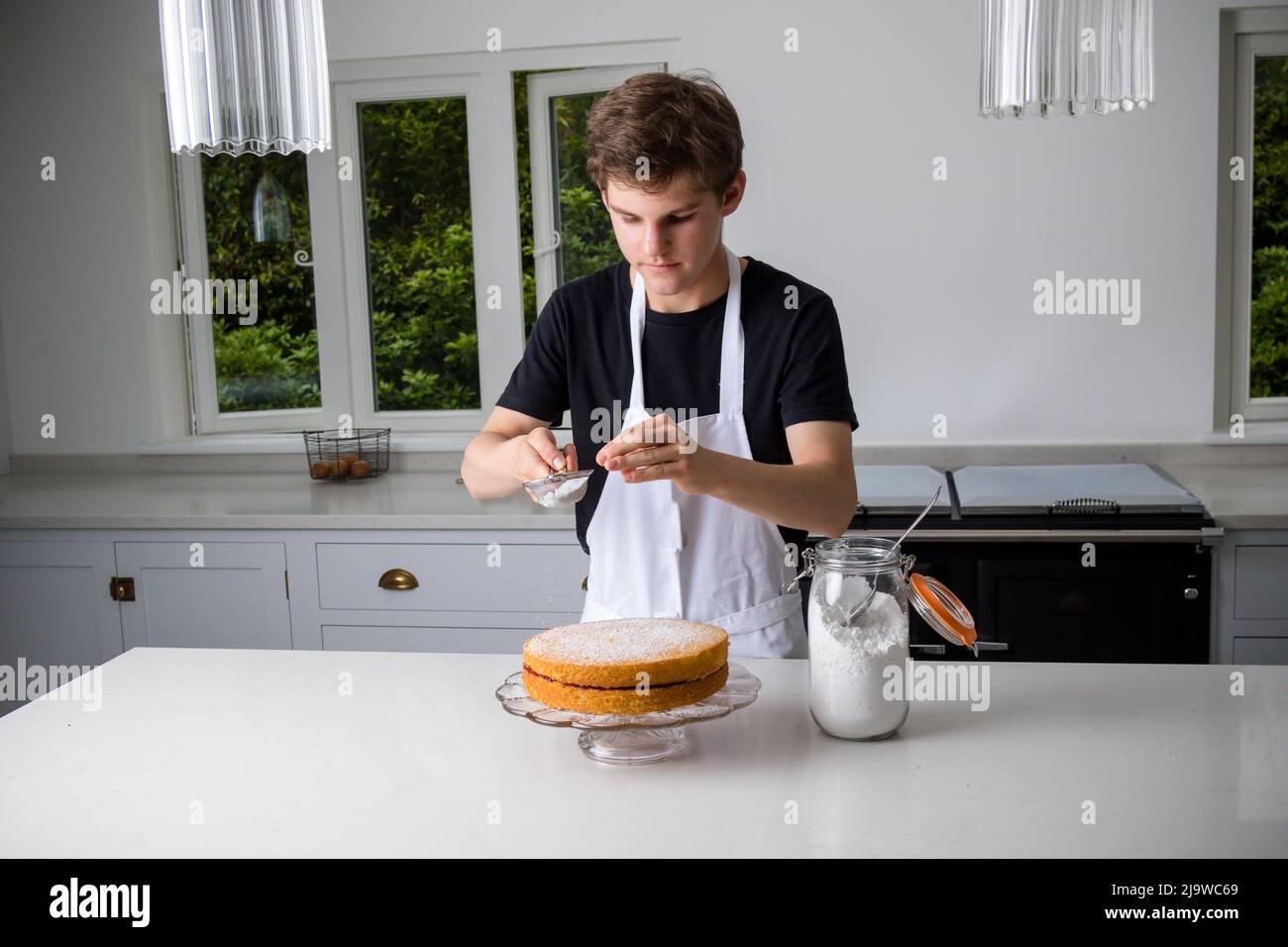 A teenage boy in a domestic kitchen putting icing sugar on a cake Stock ...