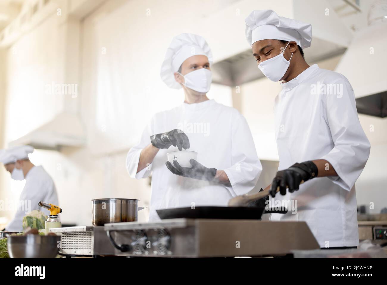 Multiracial team of cooks in uniform and face masks cooking meals for a ...