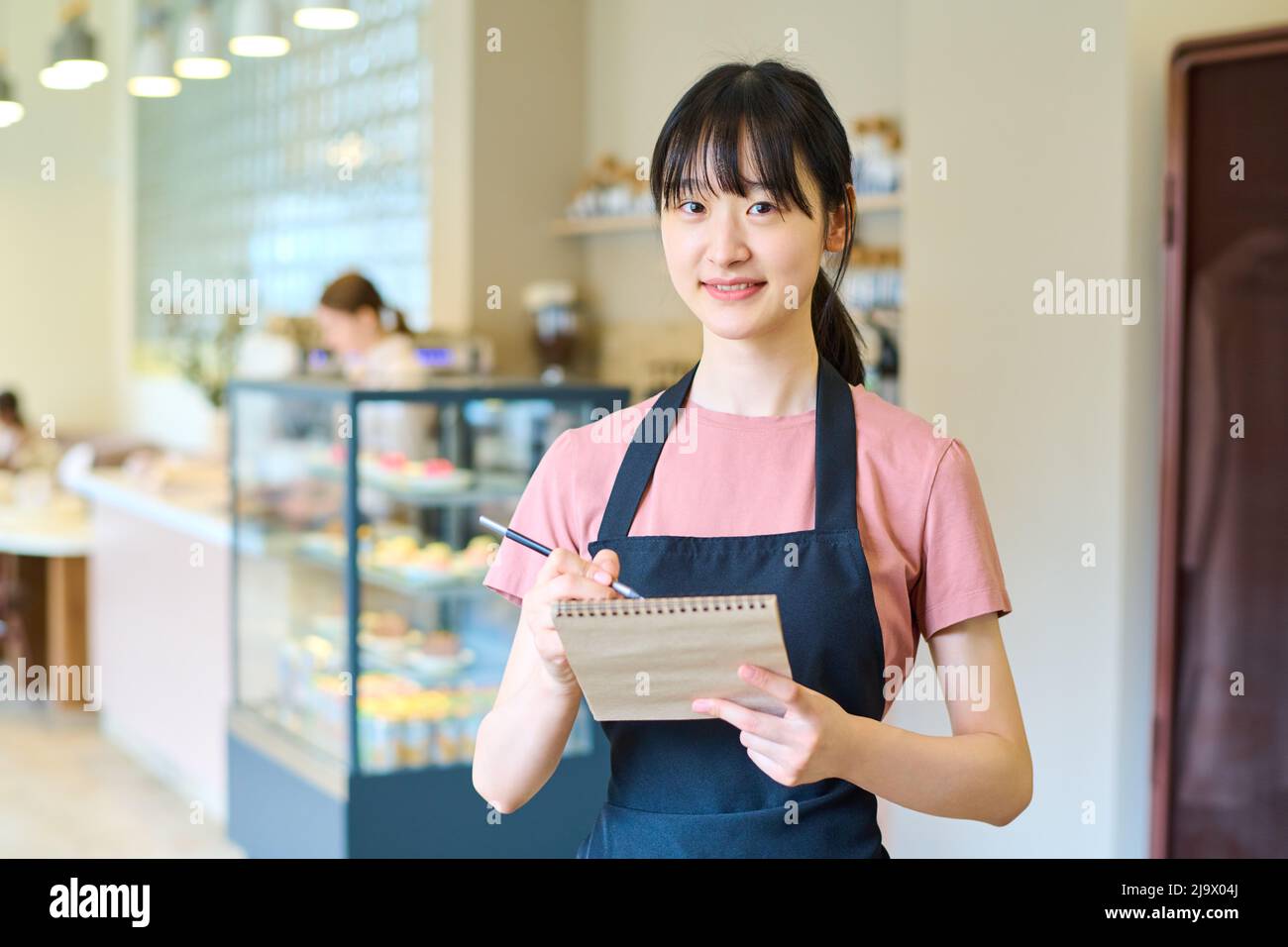 Portrait of Asian young woman in apron making notes in notepad while ...