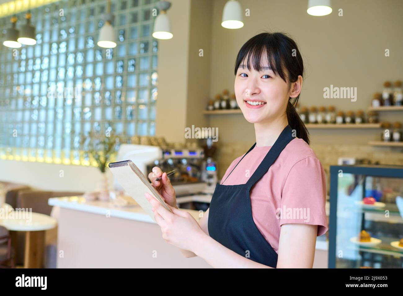Portrait of friendly Asian waitress in black apron standing with ...