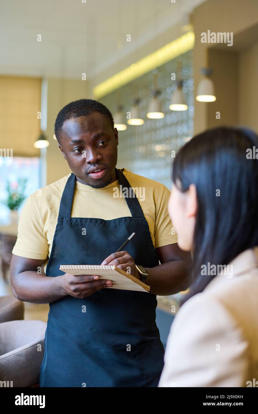 African young waiter in apron talking to woman in cafe and writing her ...