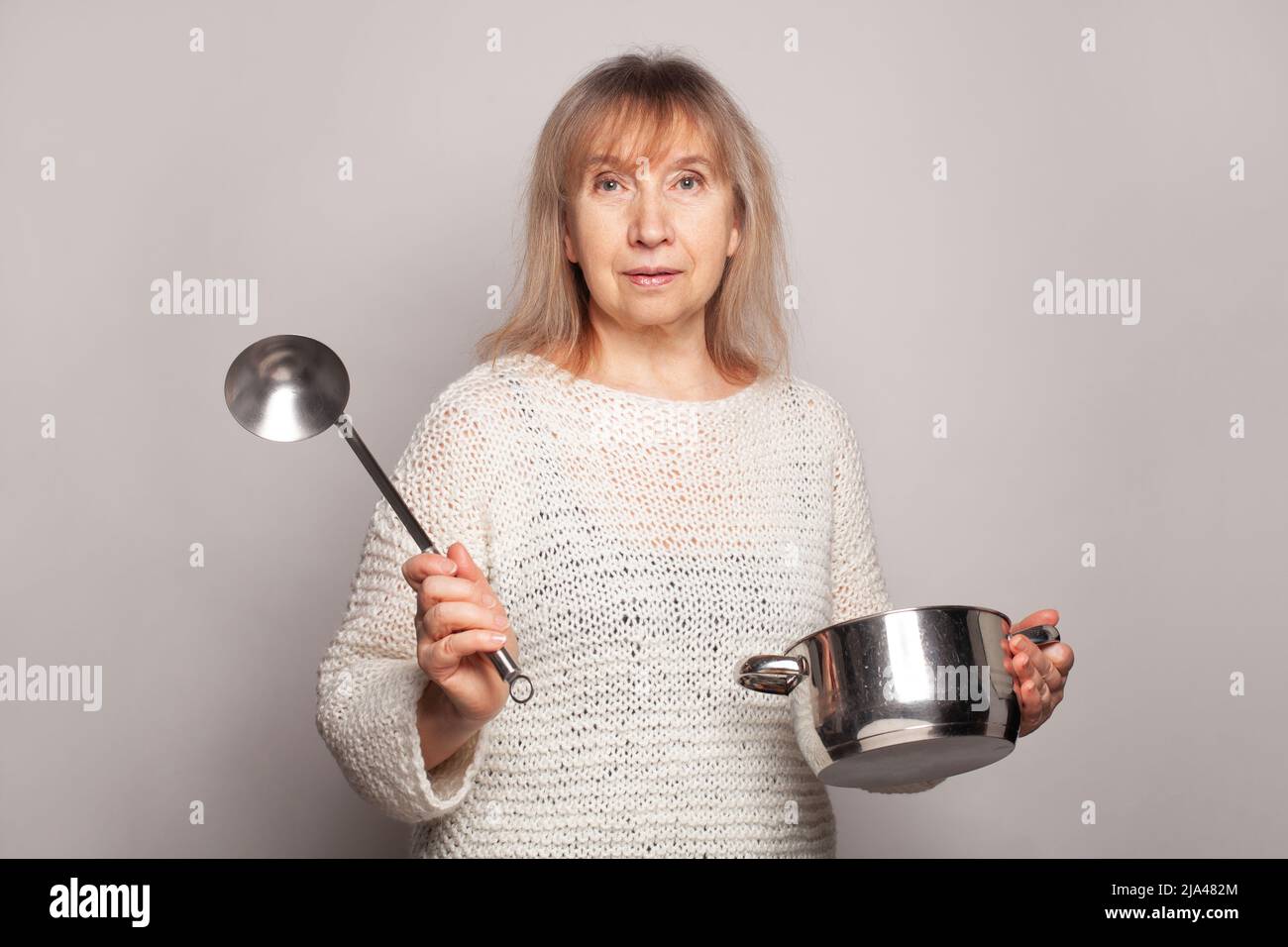 Portrait of senior woman with ladle and saucepan on white studio wall ...