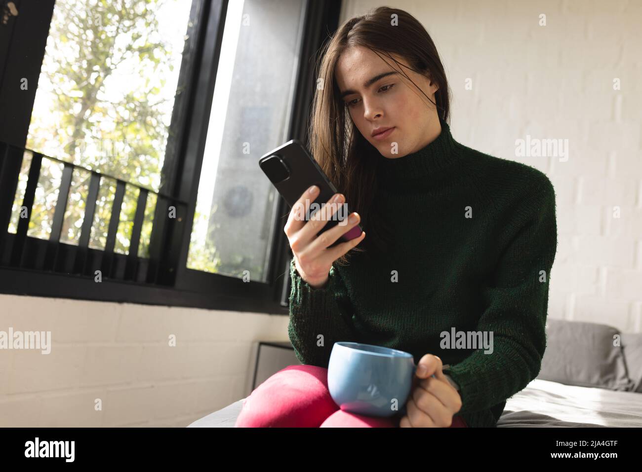 Non-binary trans woman holding coffee cup using smartphone in bedroom ...
