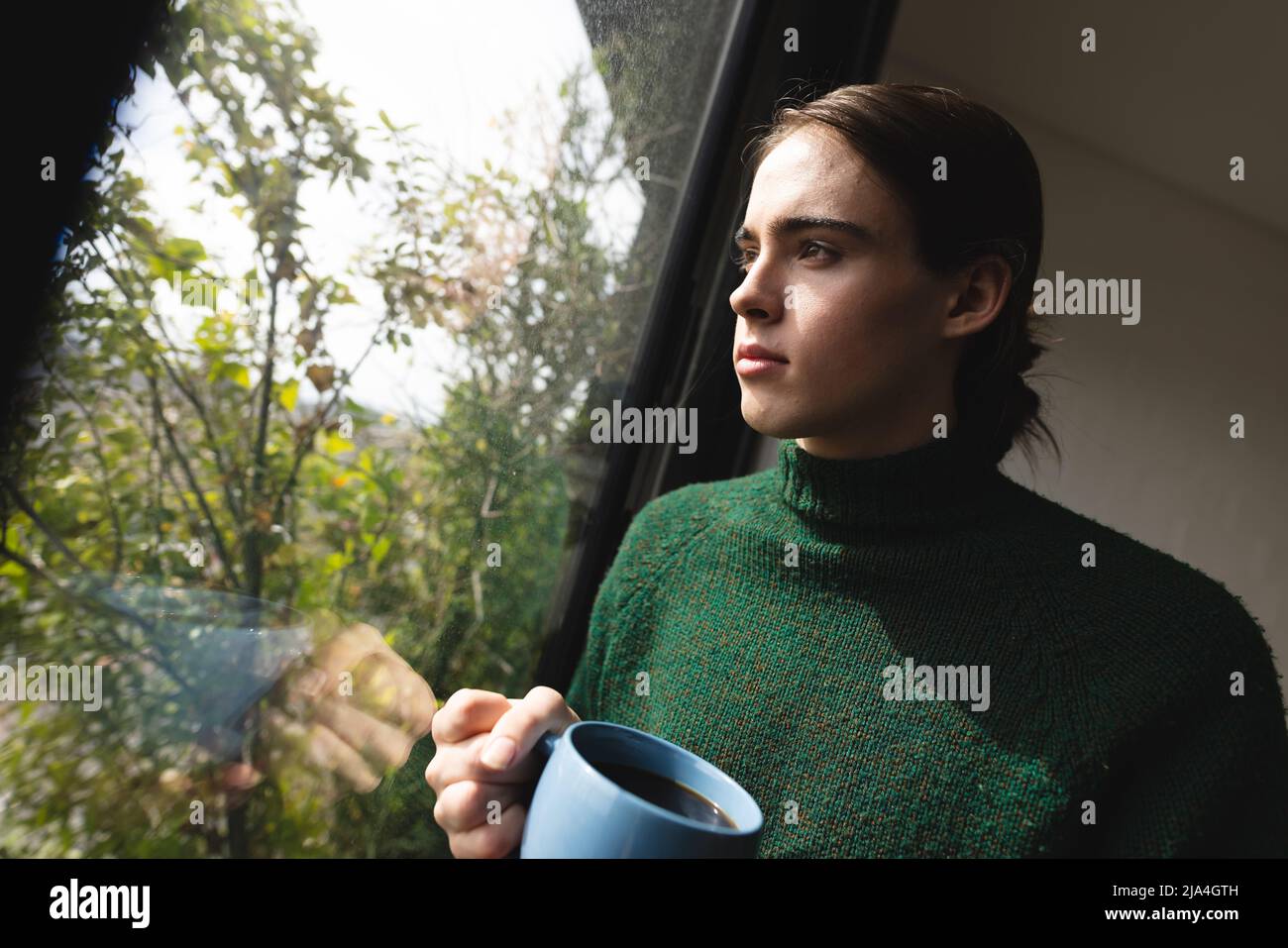 Non-binary trans woman holding coffee cup looking out of the window at ...