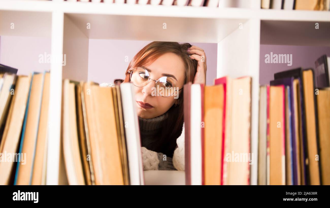 Thoughtful woman in glasses looking at books and scratching head ...