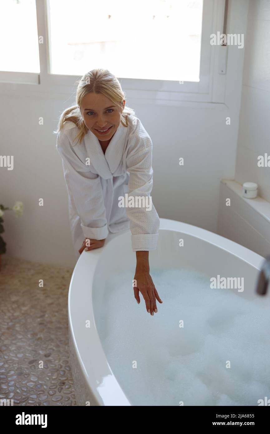Portrait of pretty blonde Caucasian woman in gown preparing bath with ...