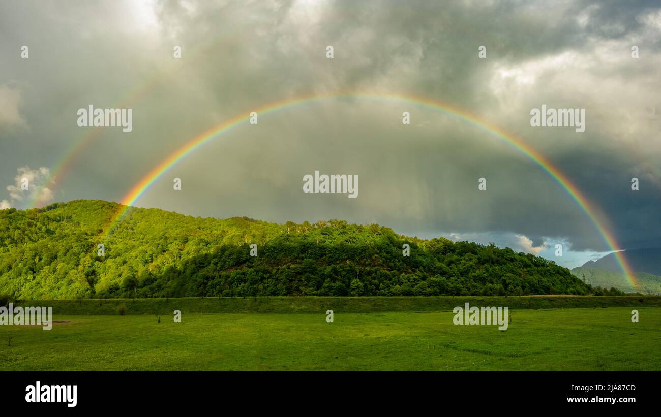 An arched, colorful rainbow appearing above a rural area during a rainy ...