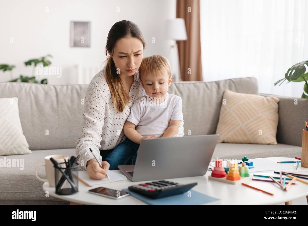 Young mother multitasking, working remotely on laptop while taking care ...