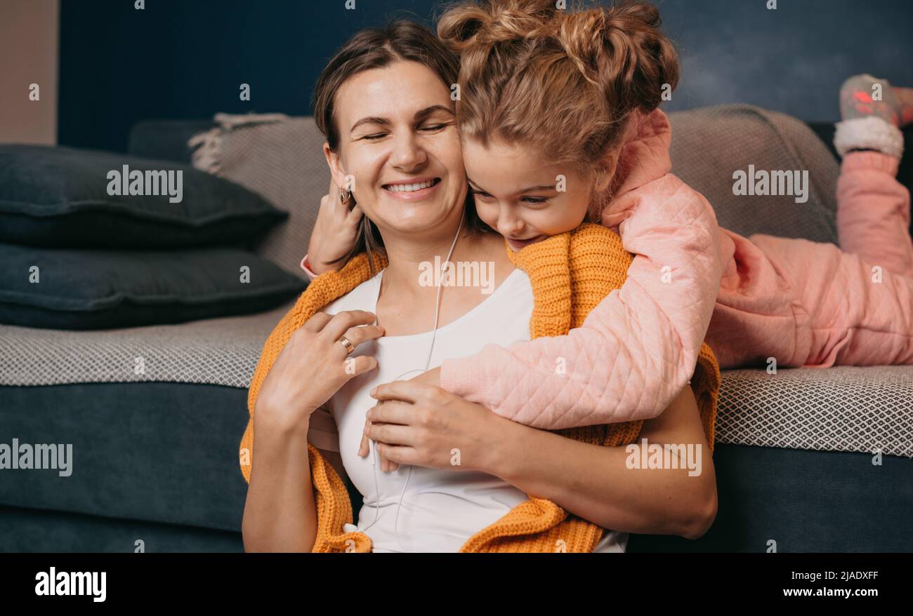 Smiling mother using laptop and earphones sitting on the floor while her daughter hugging her ...