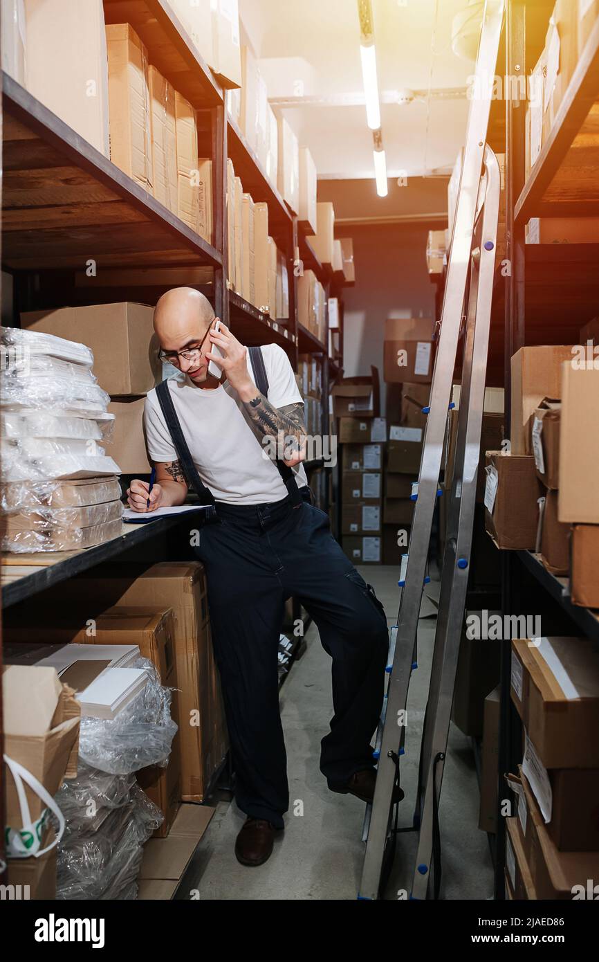 Overworked male warehouse worker talking on the phone, writing on a ...