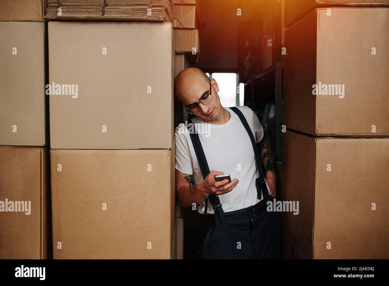 Tired male warehouse worker leaning on stack of boxes, looking at his ...