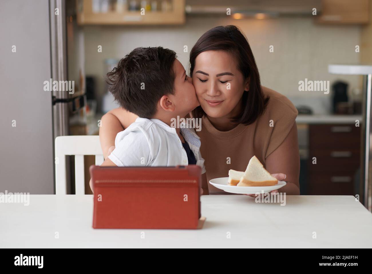 Adorable little boy kissing mother on cheeks after she brought him sandwiches for breakfast ...