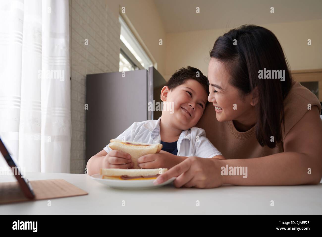 Joyful mother and son eating ham and cheese sandwiches for breakfast ...