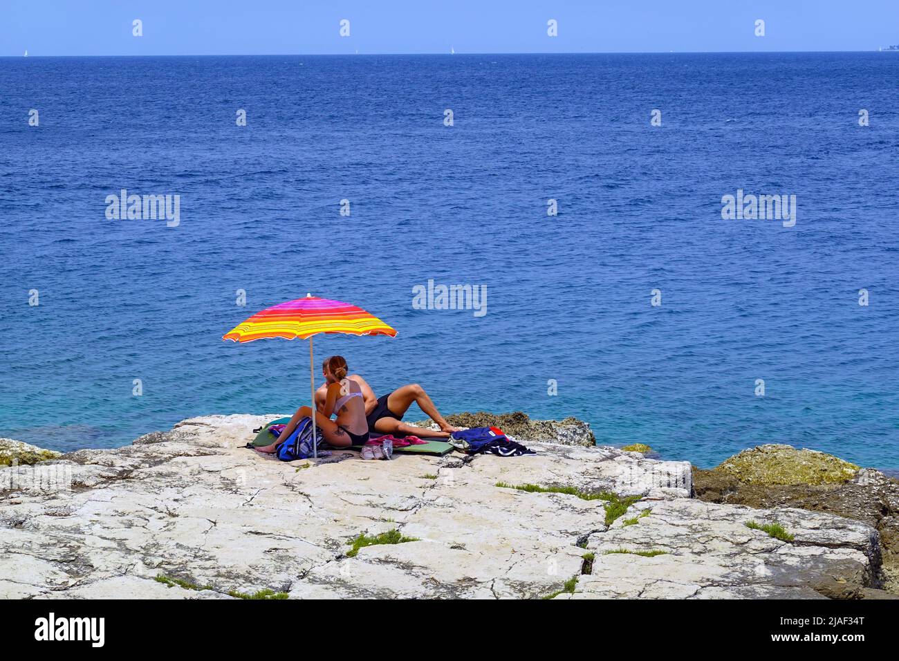 Couple sat under red and yellow beach umbrella on rocks, near thee old ...