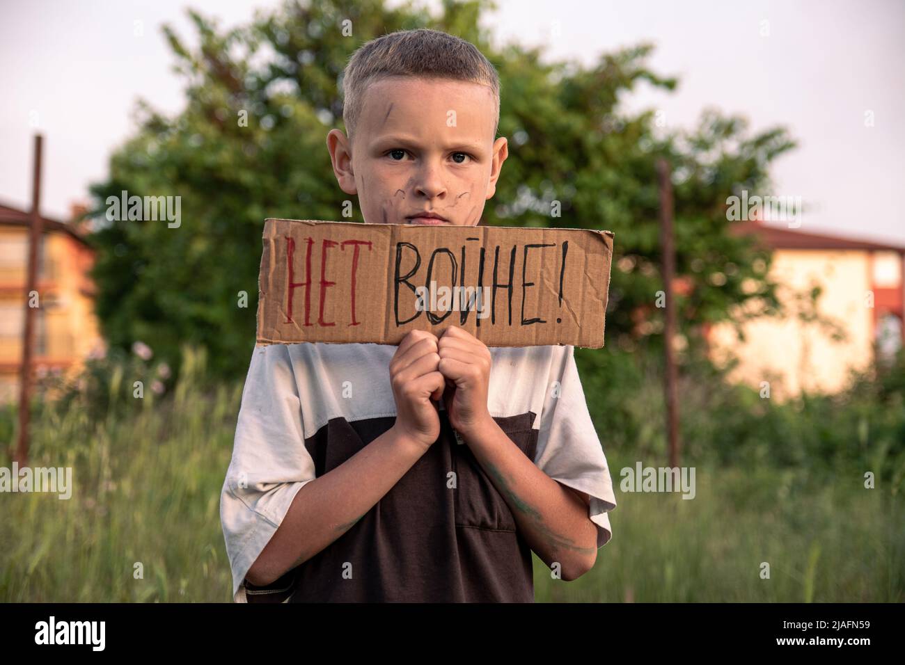 A boy with a grimy face and sad eyes holds a cardboard poster with the ...
