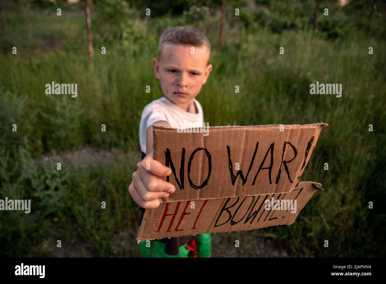 A boy with a grimy face and sad eyes holds a cardboard poster with the ...